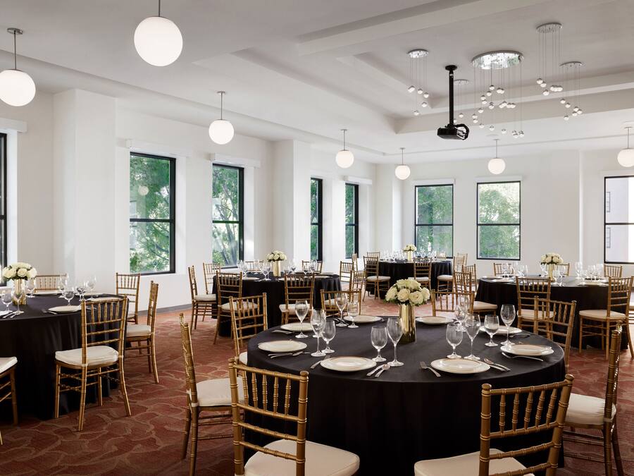 Uma ampla sala de banquetes com mesas redondas preparadas para refeições, grandes janelas e luminárias modernas.