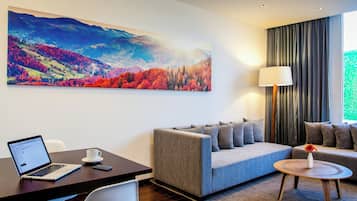 Quarto de hotel moderno com uma grande pintura de paisagem, um sofá, uma mesa de centro e uma escrivaninha com um laptop e uma xícara.