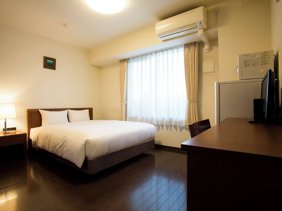 Quarto de hotel com cama, mesa de cabeceira, luminária, ar condicionado, janela com cortinas e escrivaninha com cadeira.