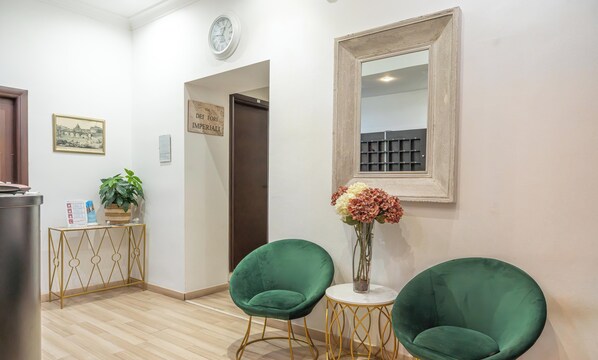 Um corredor com duas cadeiras verdes, uma pequena mesa com um vaso de flores, um relógio e um espelho emoldurado na parede.