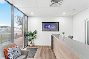 Uma área de recepção moderna com uma janela ampla, uma TV montada, um balcão de recepção, um vaso de planta e um espaço de estar com almofada estampada.