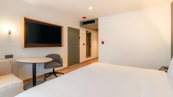 Quarto de hotel com cama, escrivaninha, cadeira, televisão e porta para o banheiro.
