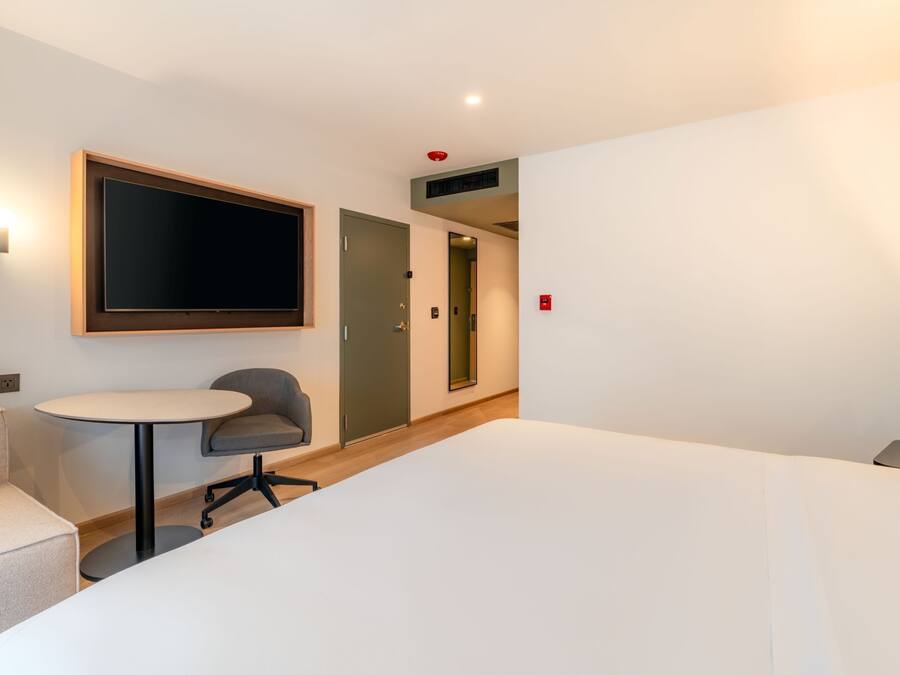 Quarto de hotel com cama, escrivaninha, cadeira, televisão e porta para o banheiro.
