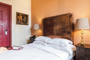 Quarto de hotel com cabeceira de madeira, roupa de cama branca, porta vermelha, um quadro na parede e duas luminárias de cabeceira.