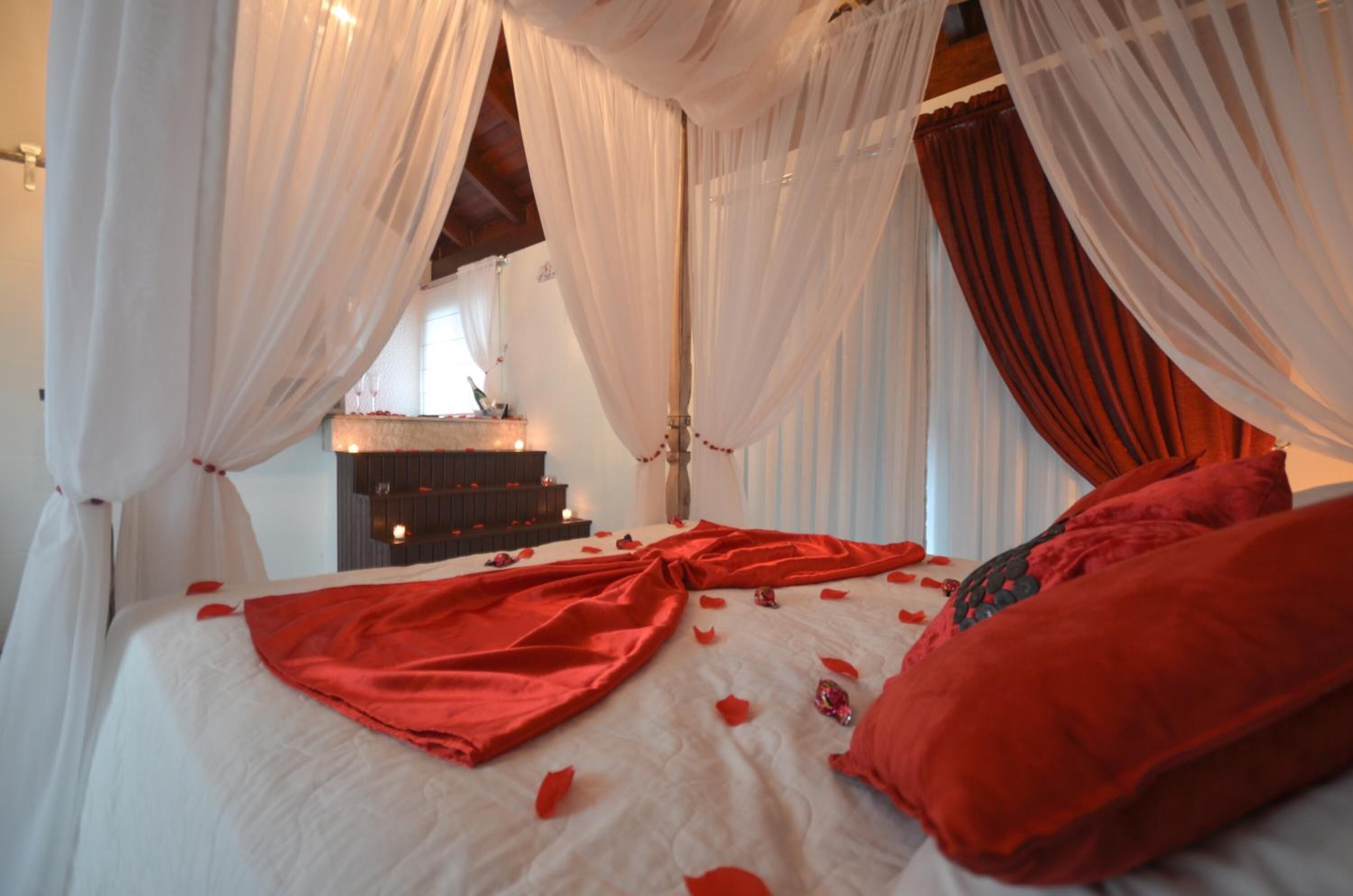 Uma cama com roupa de cama branca e vermelha, um dossel e pétalas de rosa espalhadas sobre a cama.