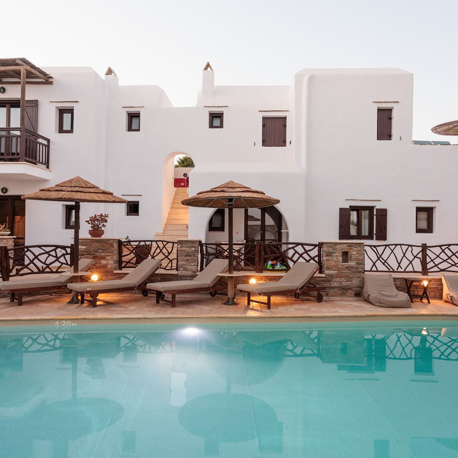 Uma área junto à piscina com espreguiçadeiras e guarda-sóis, um muro de pedra e um edifício branco com varandas.
