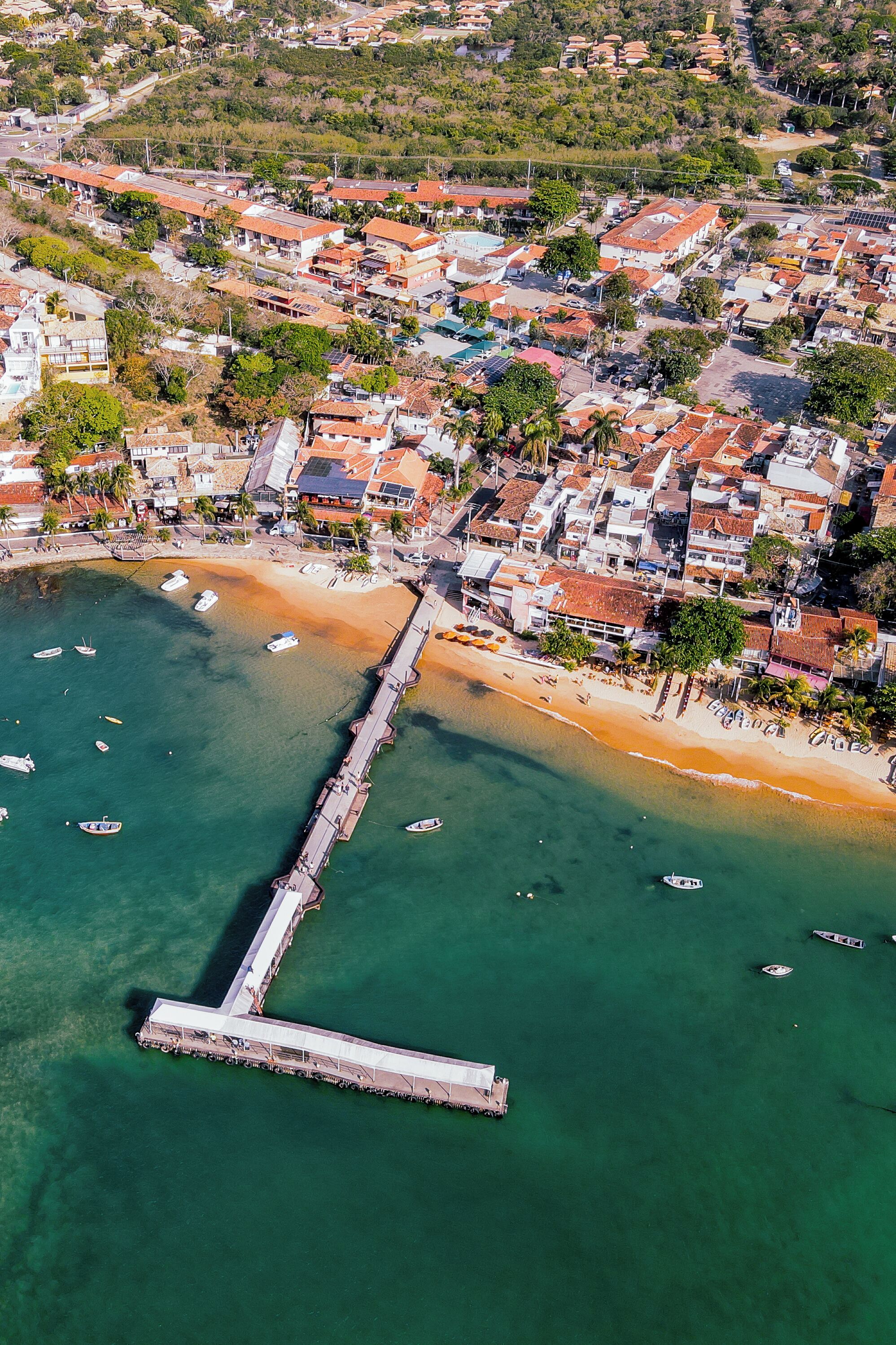 Vista aérea de uma vila costeira com um cais que se estende para o mar, rodeada por edifícios residenciais e uma praia de areia.