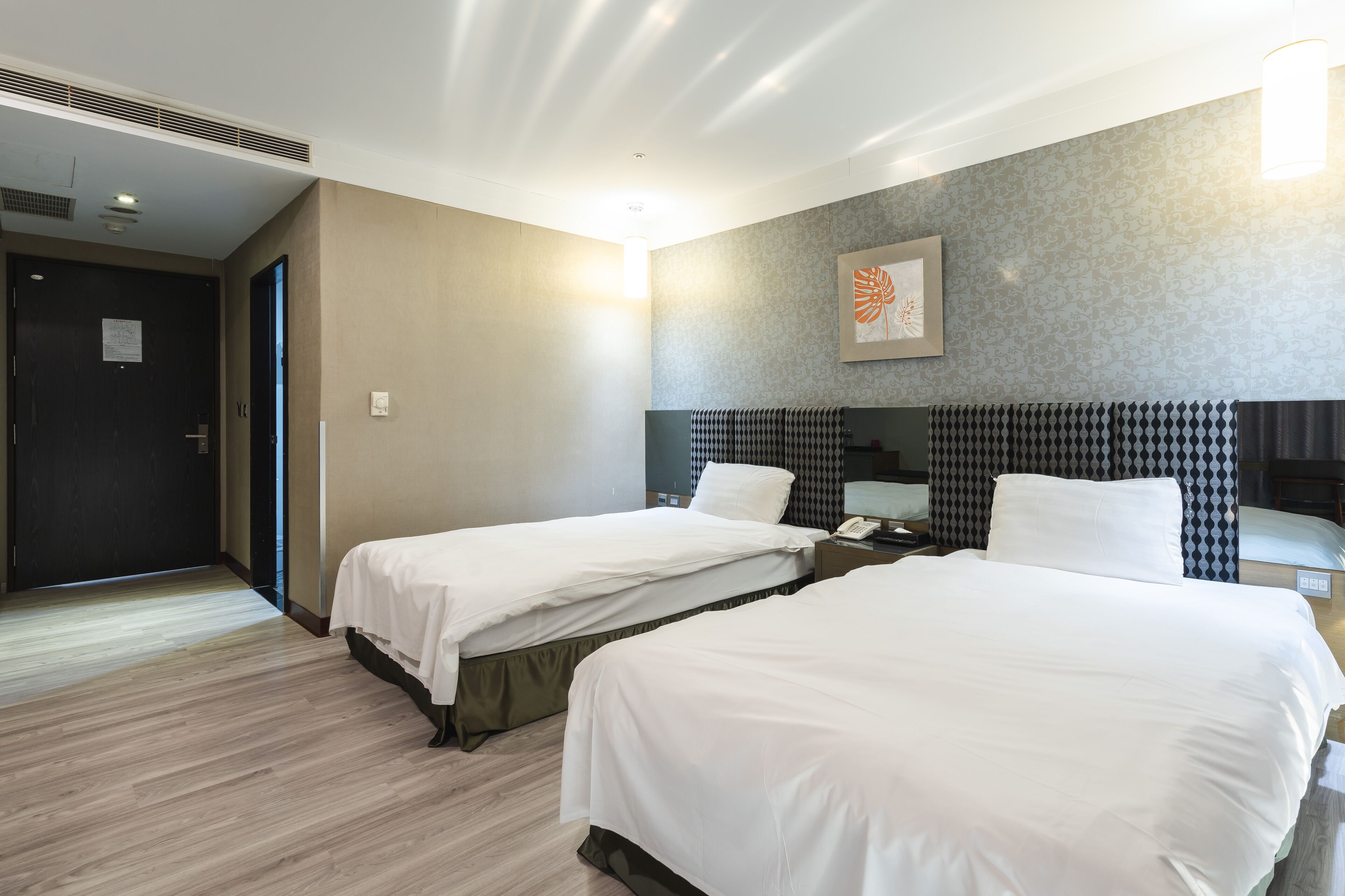 Quarto de hotel com duas camas, um painel de cabeceira estampado, uma obra de arte emoldurada e uma porta.