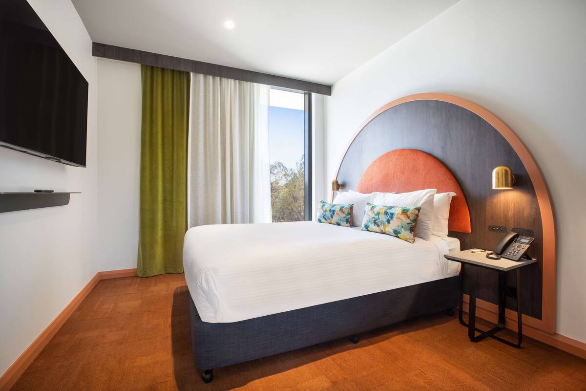Quarto de hotel moderno com uma cama grande, uma escrivaninha com telefone, uma TV fixada na parede e uma janela com cortinas.