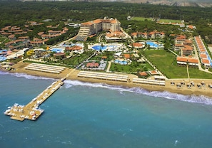 Vista aérea de um resort costeiro com vários edifícios, um cais que se estende para o mar e uma área de praia com espreguiçadeiras.