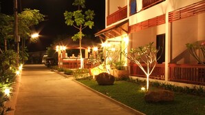 Vista noturna de um hotel com um caminho pavimentado, área de estar externa e paisagismo bem cuidado.