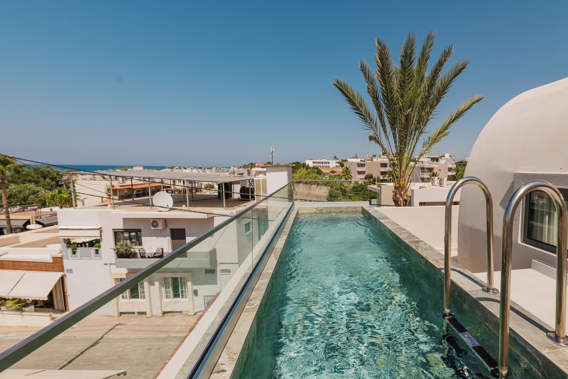 Piscina no terraço com águas cristalinas azuis, rodeada por edifícios modernos e palmeiras, com vista para a área costeira.