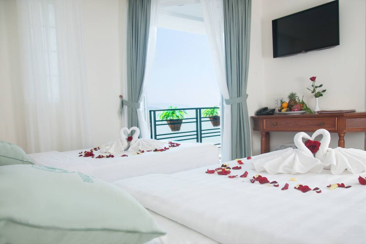 Quarto de hotel com duas camas, varanda com plantas em vasos, cómoda de madeira e televisão de tela plana.