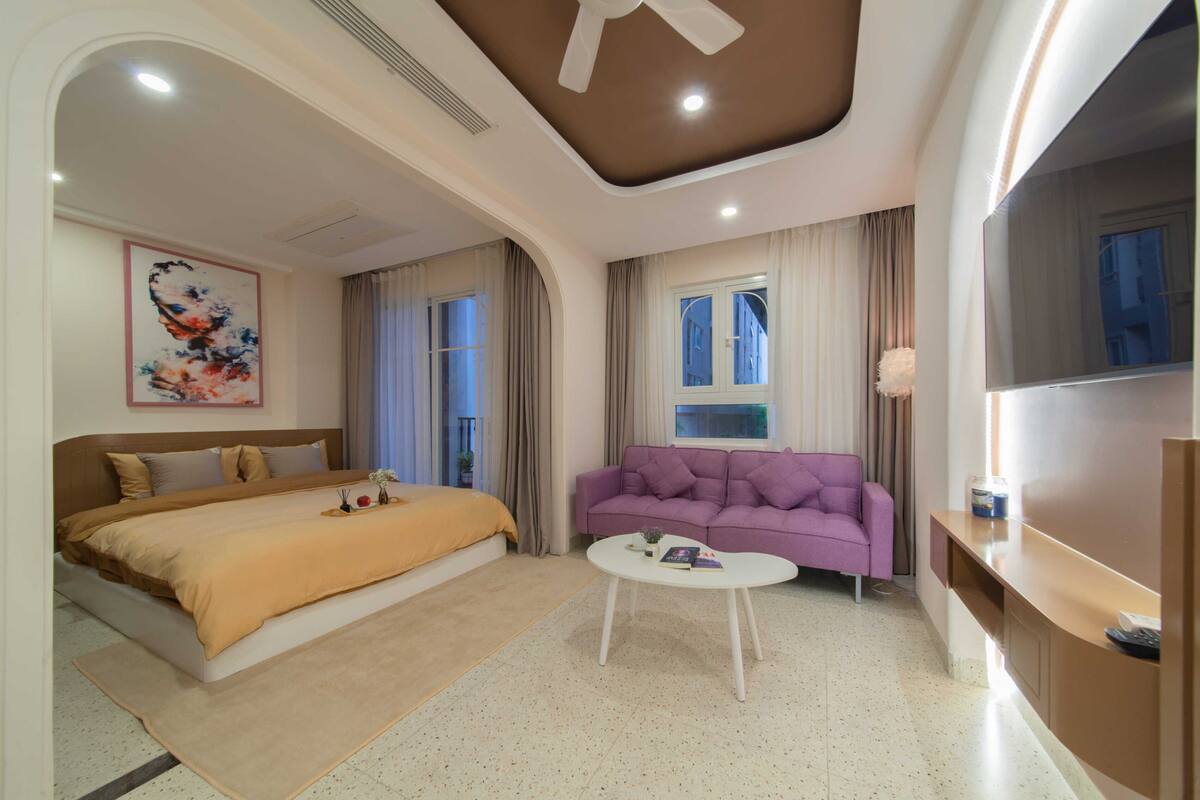 Quarto de hotel moderno com uma cama grande, um sofá roxo, uma mesa de centro branca e uma televisão fixada na parede.