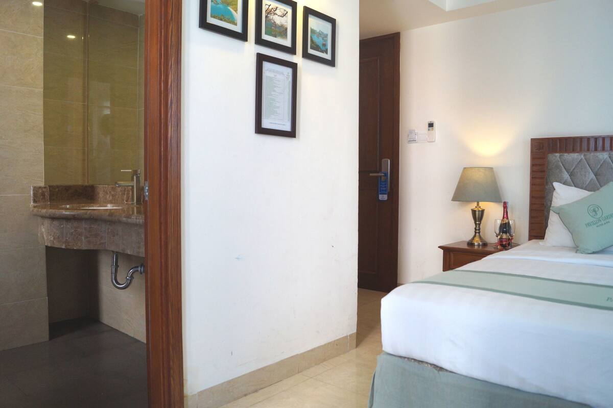 Quarto de hotel com cama, mesa de cabeceira com abajur, certificado emoldurado na parede e banheiro com pia e espelho.