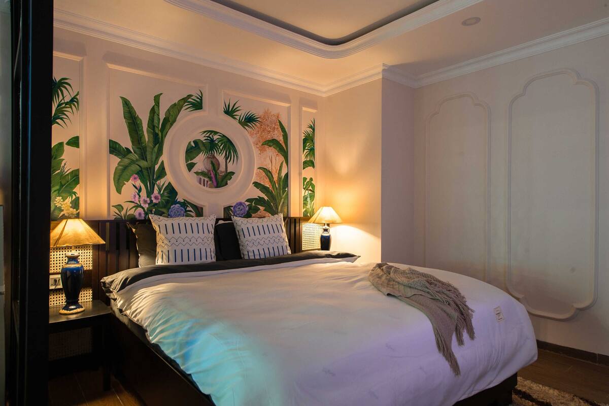 Quarto bem iluminado com uma cama grande, duas mesas de cabeceira com abajures e quadros com tema tropical.