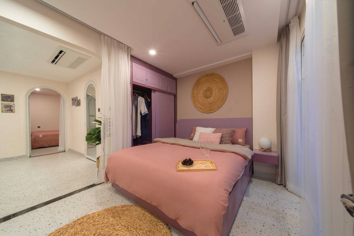 Quarto com colcha rosa, mesa de cabeceira com bandeja de frutas e um pendurar tecido na parede.