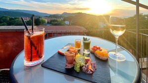 Uma mesa com um copo de vinho branco, um copo de chá gelado e diversos petiscos, numa varanda com vista para uma paisagem deslumbrante ao pôr do sol.