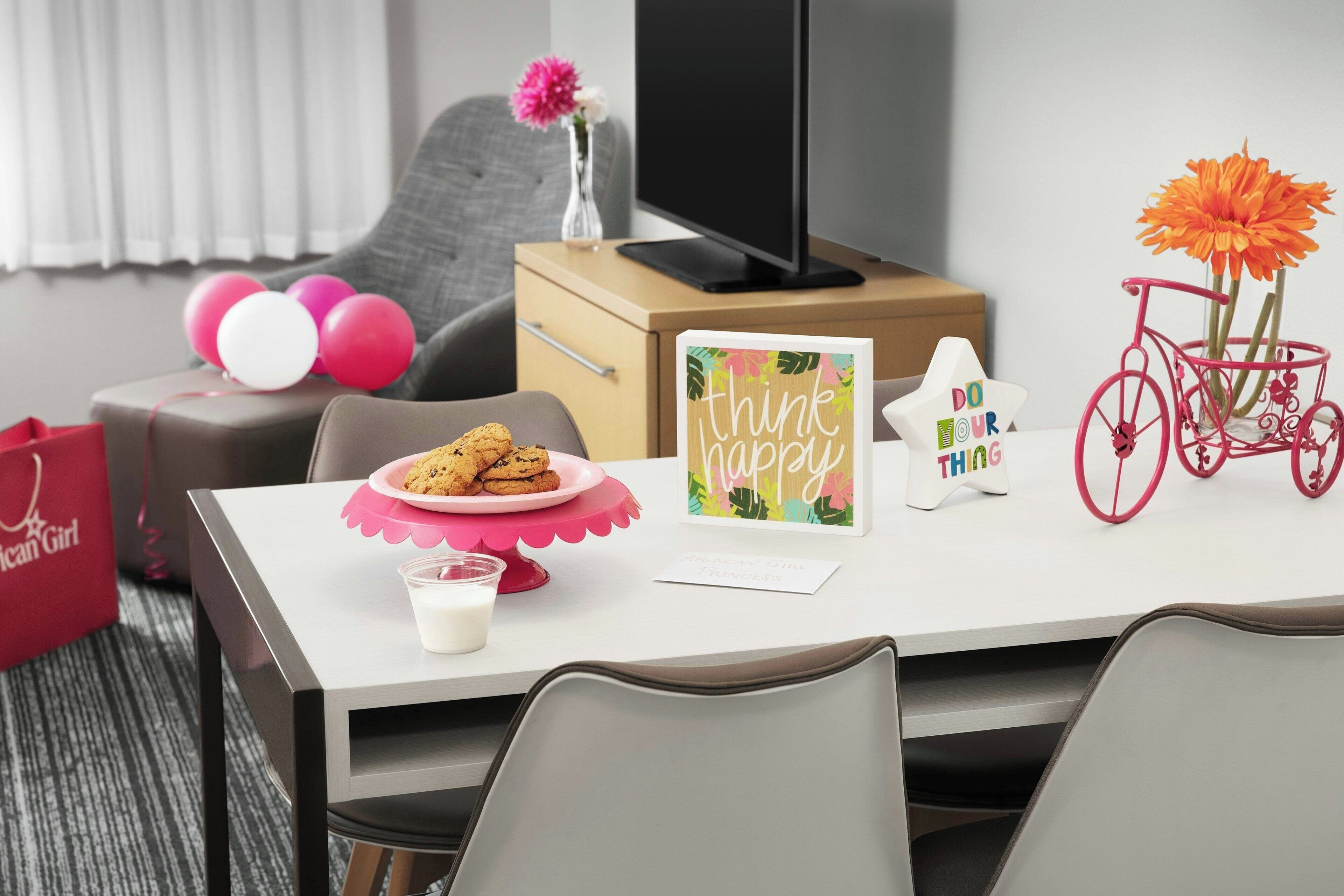Quarto de hotel com mesa de jantar posta com biscoitos e leite, televisão, vaso com flores e decoração de bicicleta.