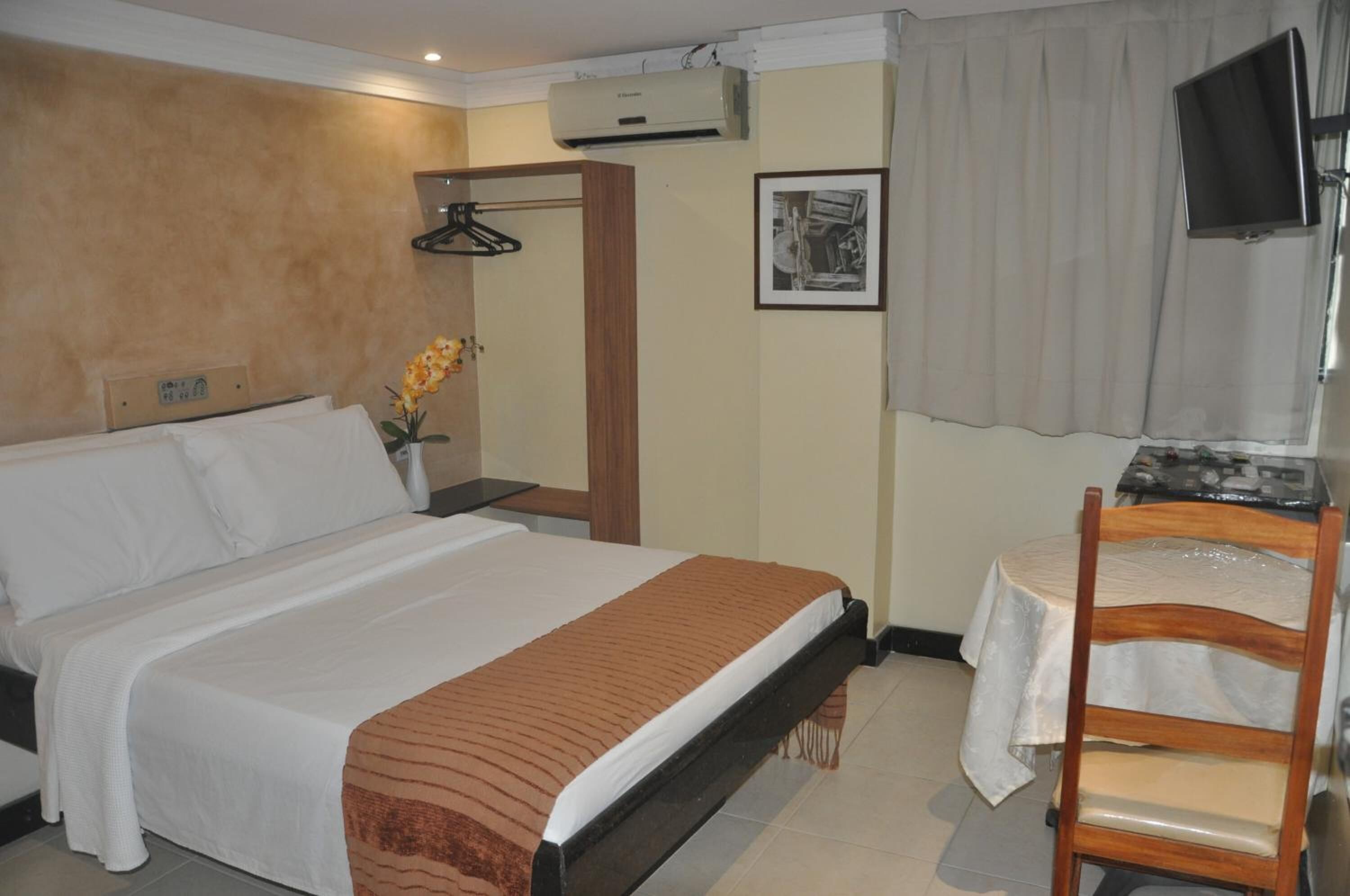 Quarto de hotel com cama, escrivaninha, cadeira, televisão e um quadro na parede.