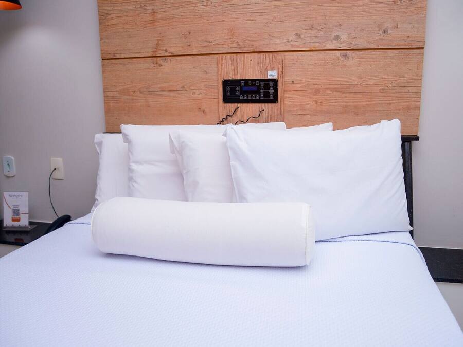 Uma cama bem arrumada com lençóis e almofadas brancas, uma cabeceira de madeira com um painel de controle e uma mesa de cabeceira com uma luminária e um telefone.