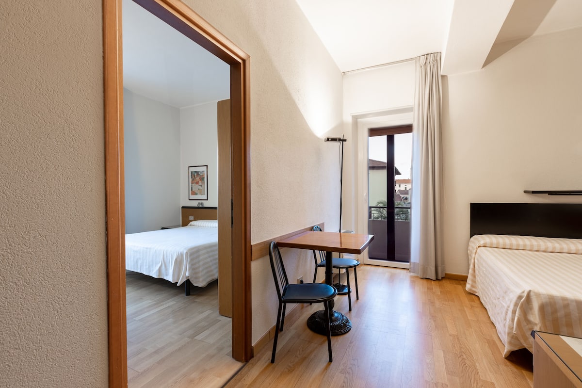 Quarto de hotel com piso de madeira, uma cama, uma escrivaninha com cadeira e vista para o exterior pela janela.