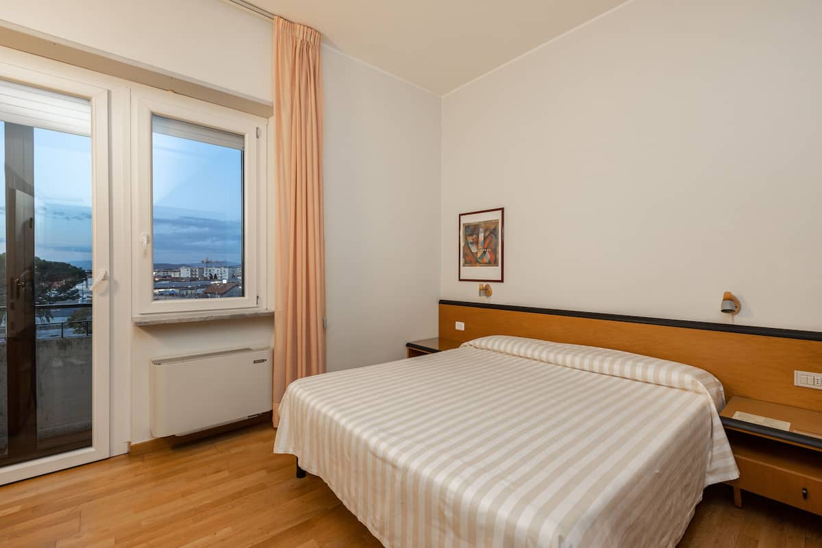Quarto com cama, mesa de cabeceira, janela com cortinas, radiador e luminária fixa na parede.