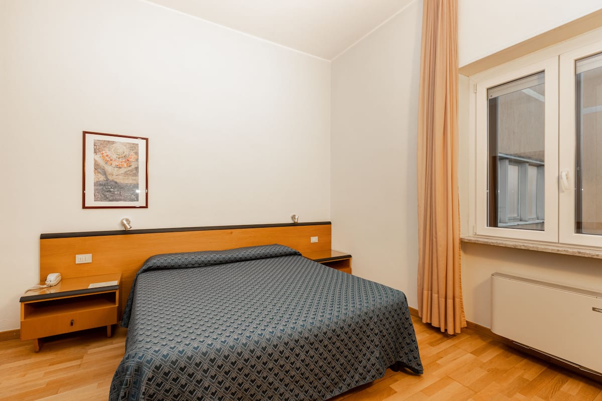 Quarto com cama, mesas de cabeceira, um radiador e uma janela com cortinas, além de um quadro na parede.