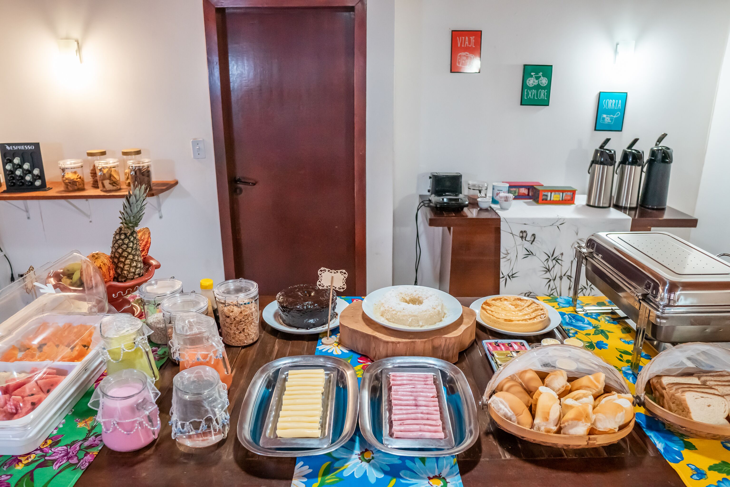 Disposição de buffet com diversos alimentos e bebidas, incluindo frutas, doces e uma estação de café.