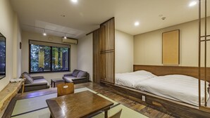 Quarto de hotel moderno com uma cama grande, uma mesa de madeira, um sofá e vista para árvores pela janela.