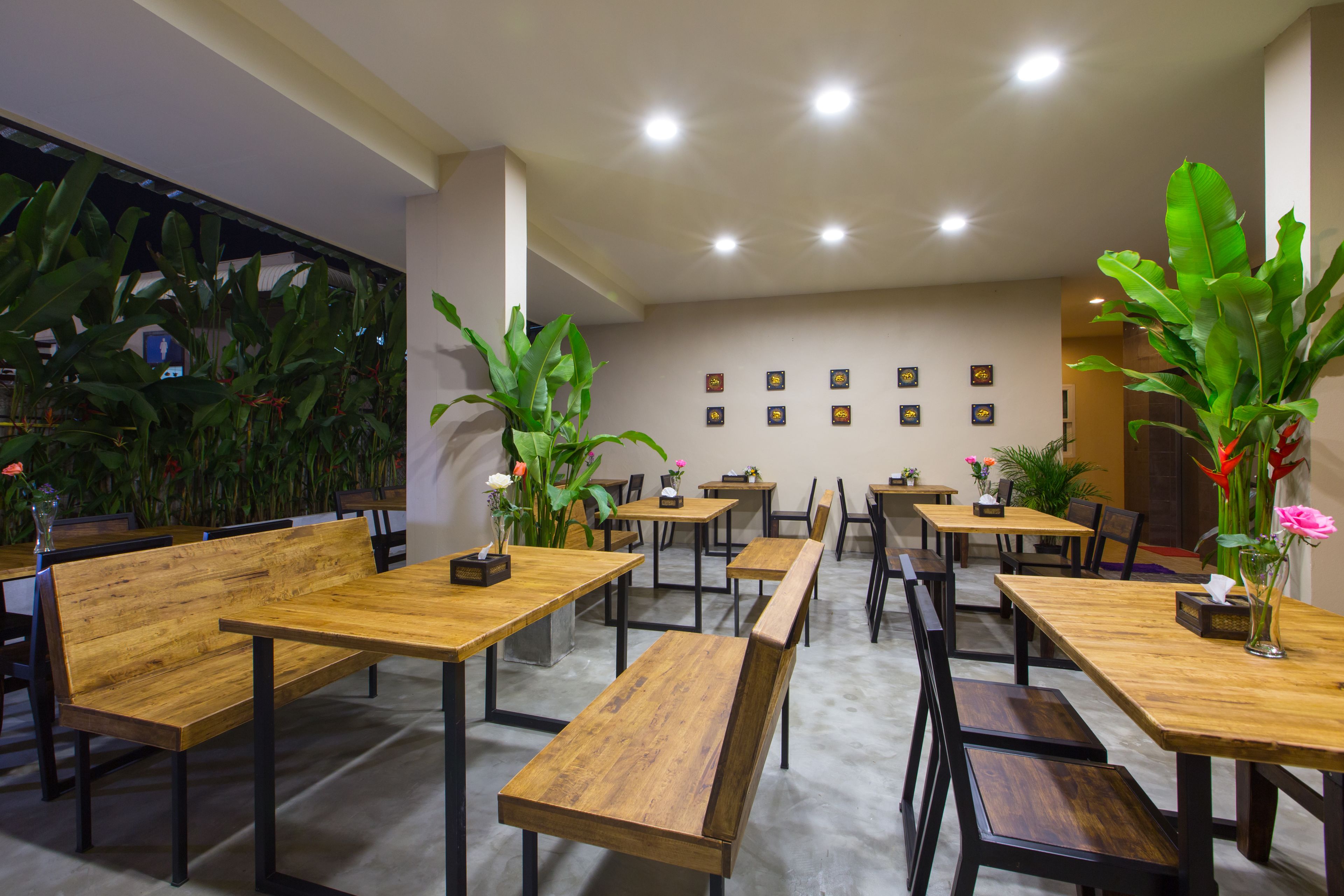 Restaurante com mesas e bancos de madeira, plantas em vasos e quadros nas paredes.