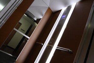 Interior de elevador com corrimão metálico, painel de comandos com botões e porta de vidro.