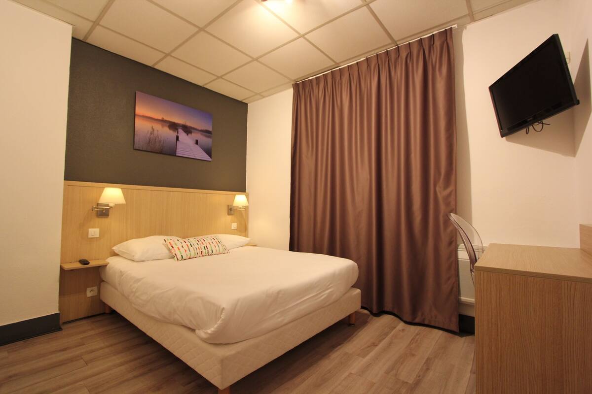 Quarto de hotel com uma cama, uma escrivaninha, uma cadeira, uma TV fixada na parede e um quadro sobre a cama.