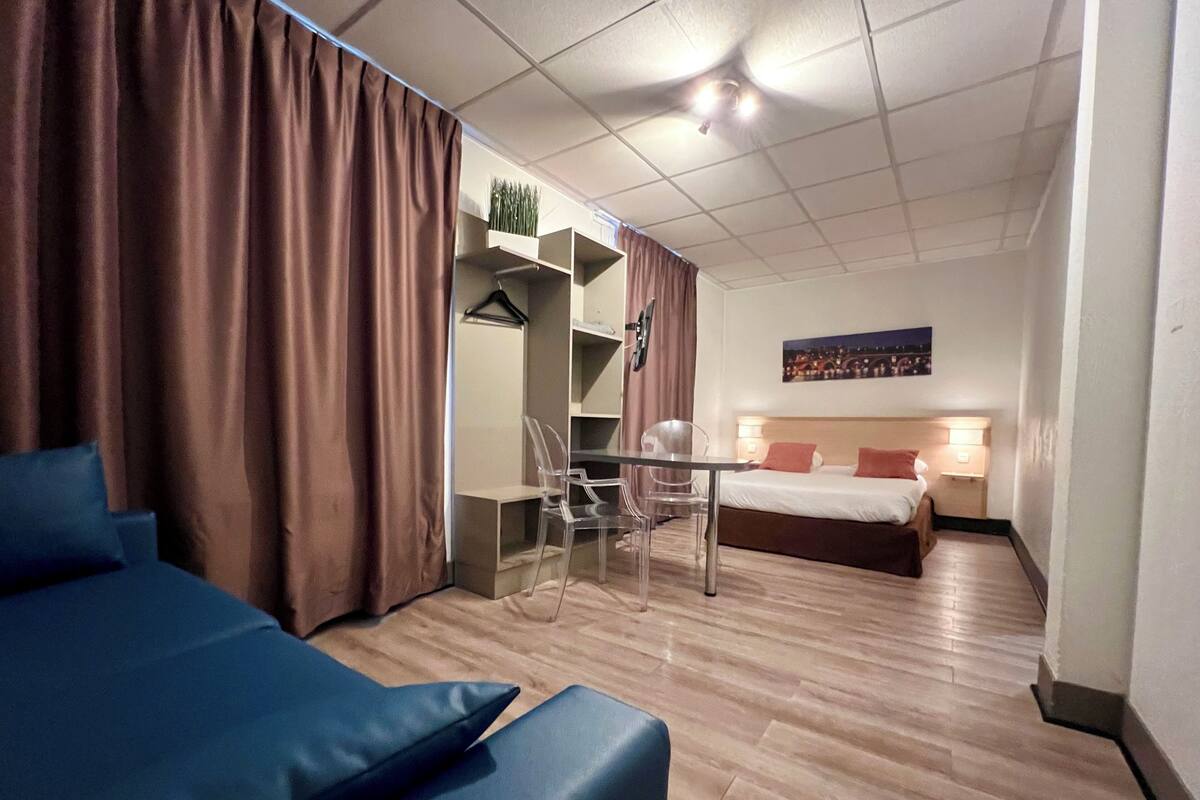 Quarto de hotel com cama, escrivaninha e cadeira. Há uma janela com cortinas, luz noturna e um quadro na parede.