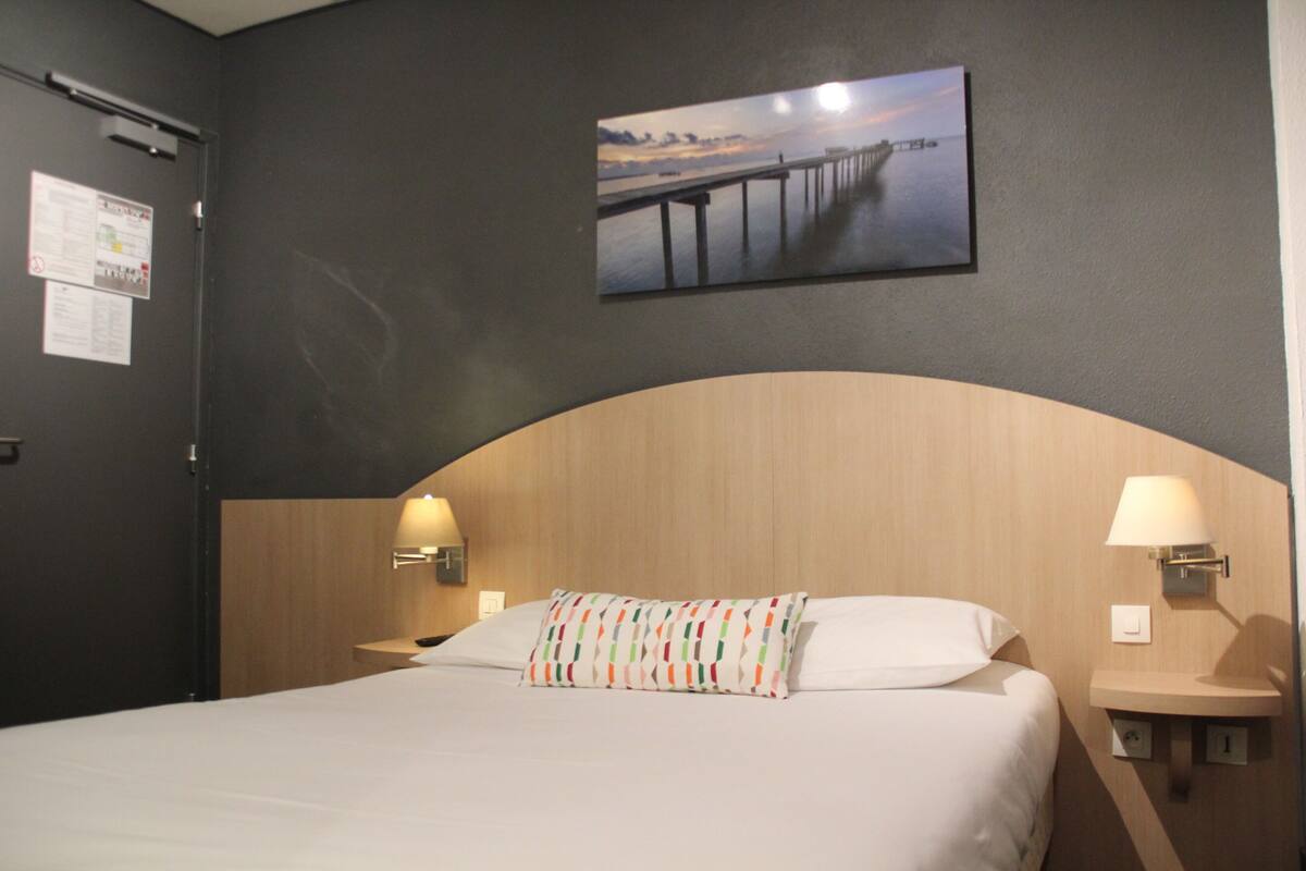 Quarto de hotel com uma cama grande, duas luminárias de cabeceira, um painel de madeira na cabeceira, uma imagem pendurada na parede de um cais e uma porta com um aviso.