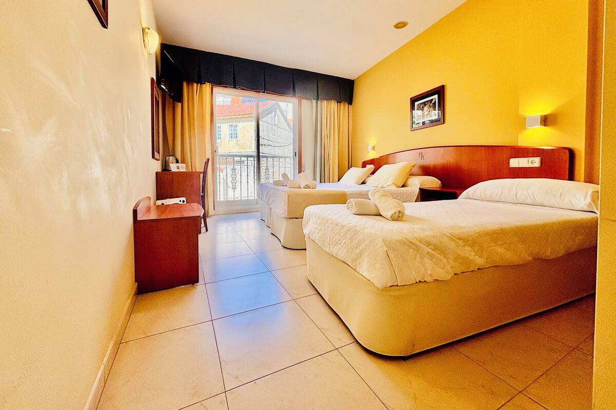 Quarto de hotel com cama, escrivaninha, cadeira, janela com cortinas e luminária fixada na parede.