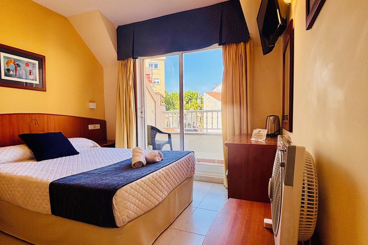 Quarto de hotel com uma cama, escrivaninha com ventilador, televisão e vista para o exterior.