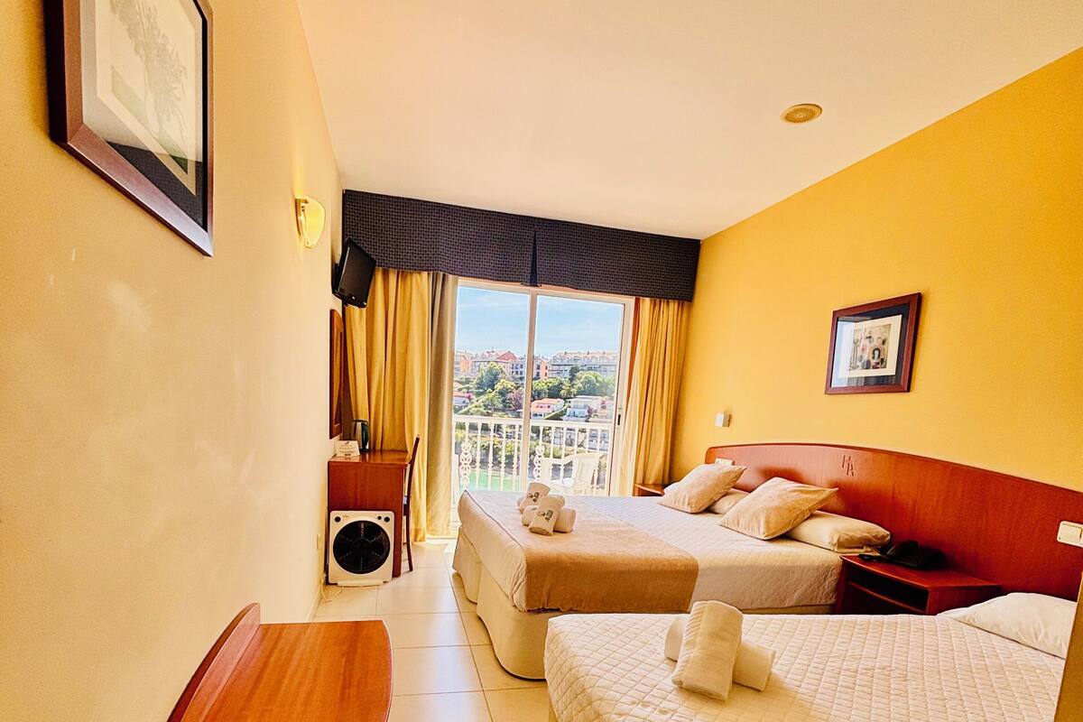 Quarto de hotel com duas camas, uma escrivaninha, uma cadeira, janela com vista e um quadro na parede.