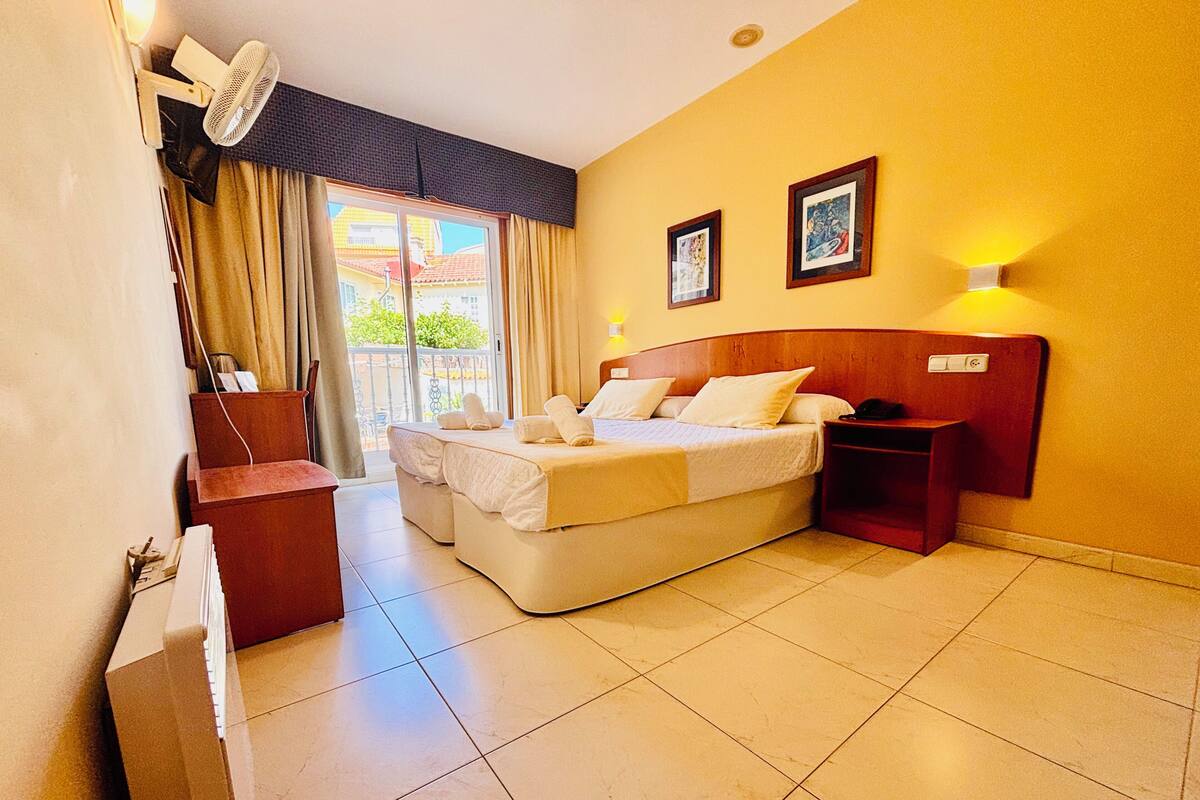 Quarto de hotel com cama, escrivaninha, cadeira, abajur e janela com cortinas.