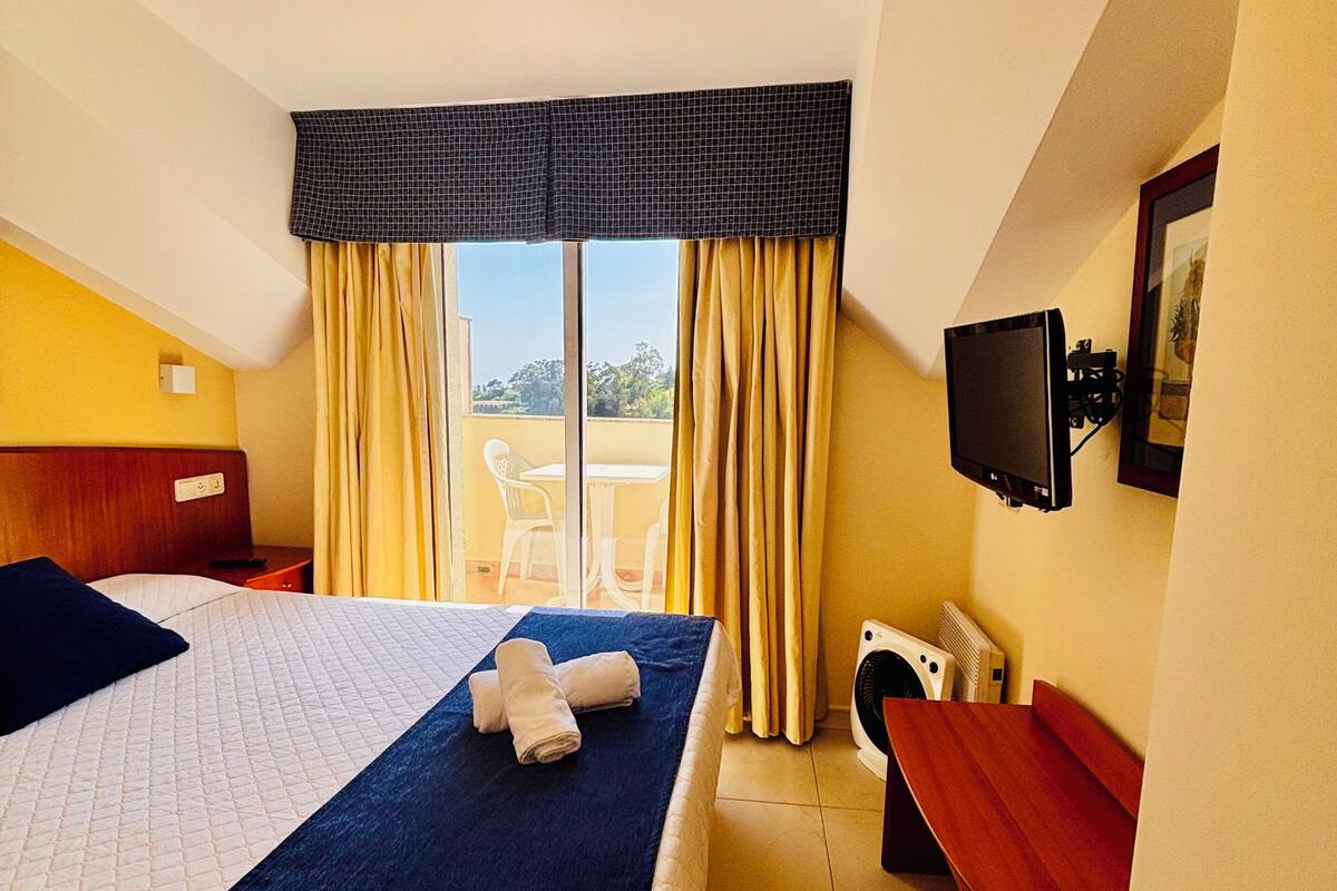 Quarto de hotel com cama, escrivaninha, cadeira, televisão e janela com cortinas.