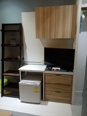 Quarto compacto com escrivaninha branca, mini-geladeira, televisão e armário de madeira.