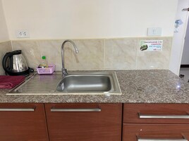 Cozinha com pia de aço inoxidável, chaleira, garrafa de detergente e cesta de alho. Há uma placa na parede e uma tomada acima da pia.