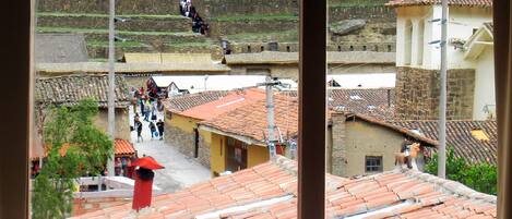 Vista através de uma janela em arco de um edifício com telhados de terracota, uma escadaria que leva a uma encosta e um pequeno edifício com telhado vermelho.