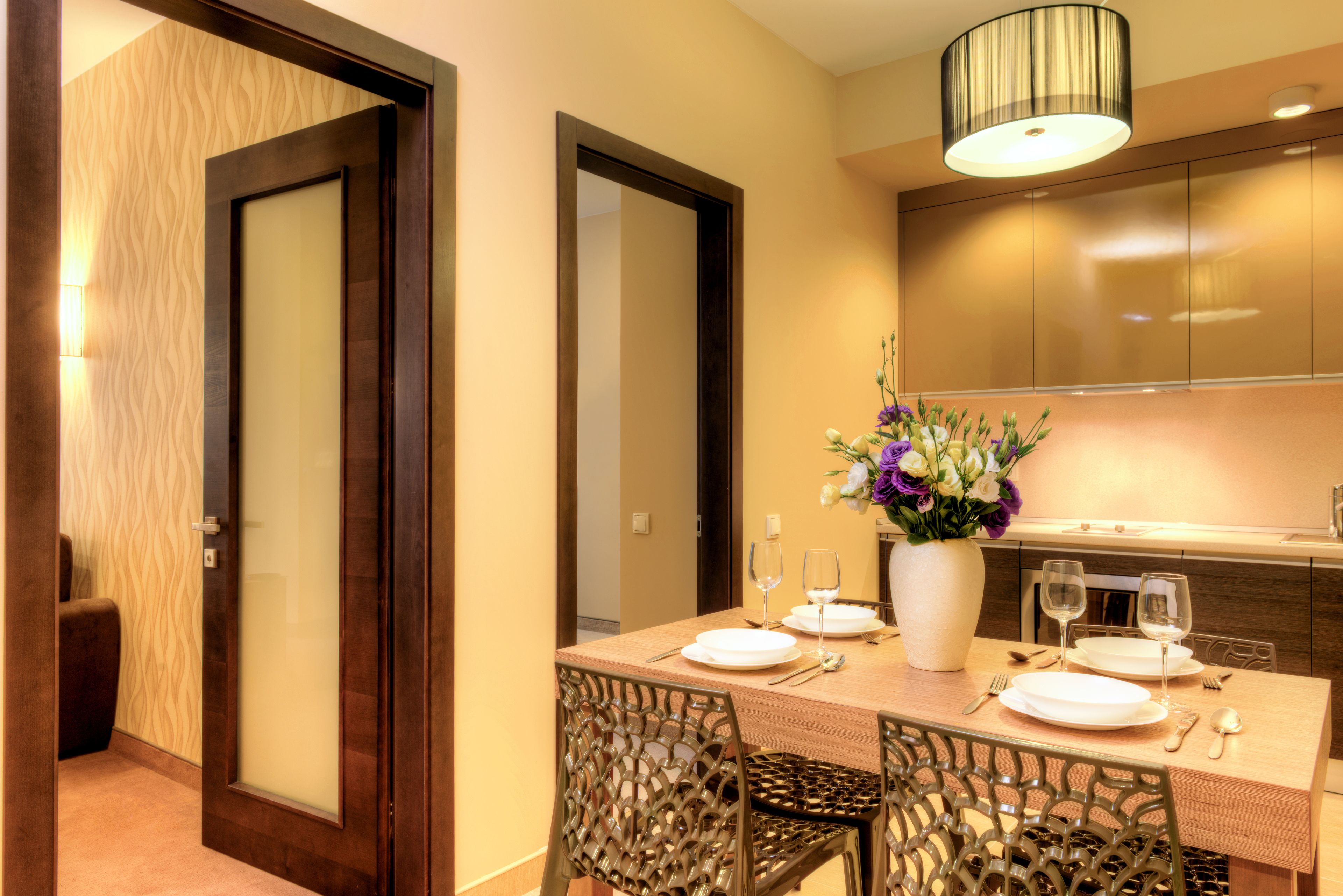 Uma sala de jantar com uma mesa de madeira posta para quatro pessoas, incluindo pratos, talheres e um vaso de flores.