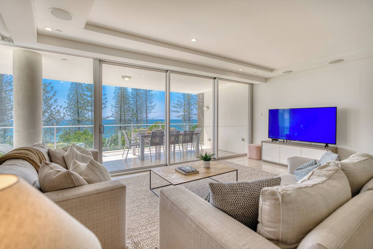 Uma sala de estar moderna com uma televisão de tela plana grande, um sofá, uma mesa de centro e vista para o exterior por meio de grandes portas de vidro.