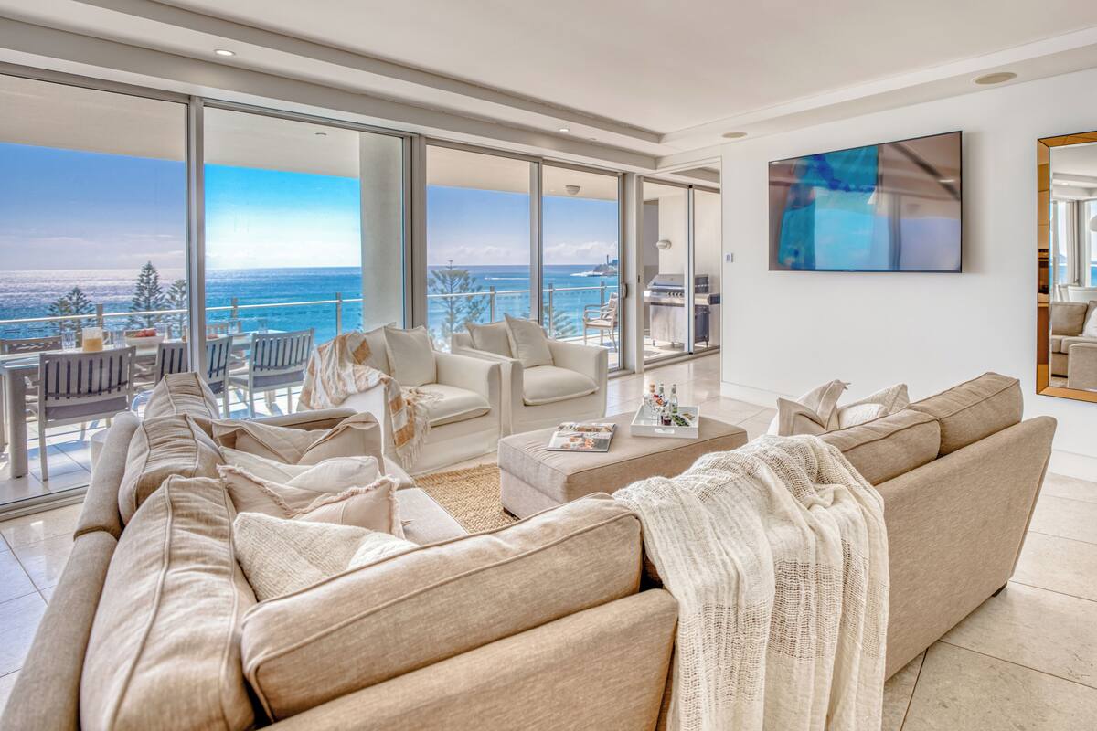 Uma sala de estar ampla com um sofá grande, uma mesa de centro e uma TV na parede. Há uma varanda com vista para o oceano e um espaço de jantar com mesa e cadeiras.