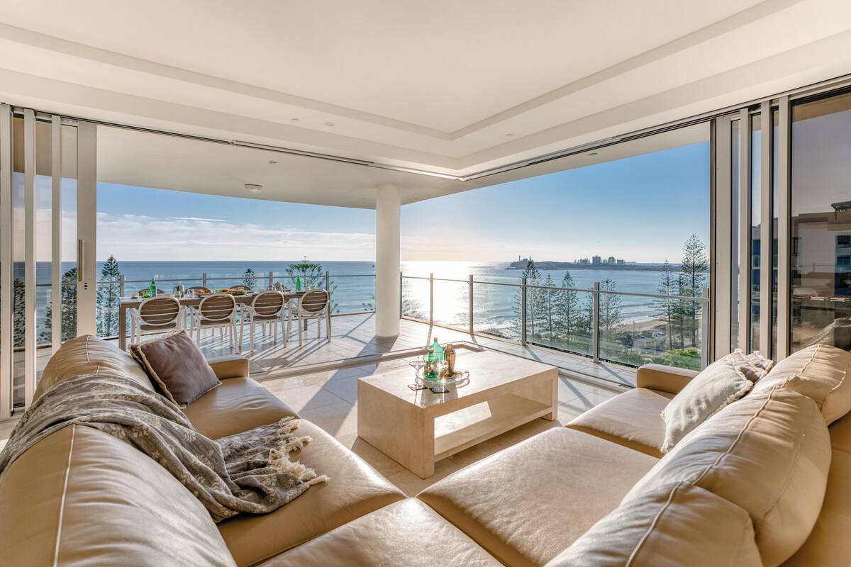 Uma sala de estar moderna com uma grande janela que oferece uma vista panorâmica do oceano, um espaço de estar aconchegante e uma mesinha com objetos decorativos.