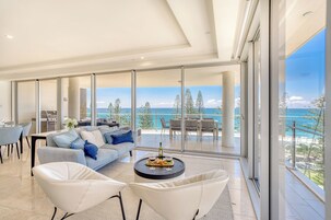 Uma sala de estar ampla com varanda com vista para o mar, mobiliada com sofá, cadeiras e mesa de jantar.