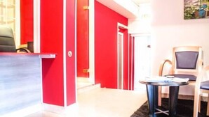 Um saguão de hotel com esquema de cores vermelho e branco, um balcão de recepção, uma área de estar com uma pequena mesa e cadeiras, e uma porta que dá para outro quarto.
