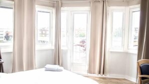 Quarto de hotel com uma cama grande, uma cadeira e vista para o exterior através de amplas janelas.